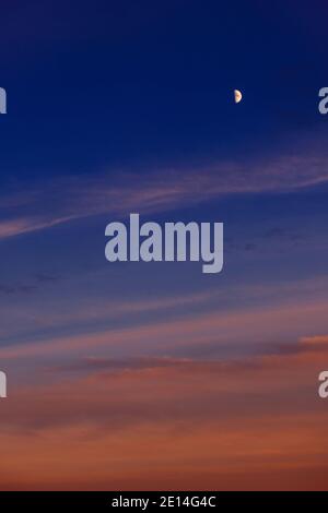 Half moon rising against deep blue sky and warm clouds as the sun sets ...