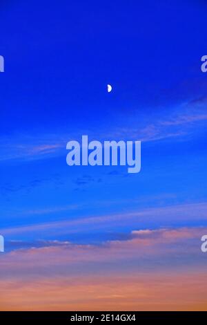 Half moon rising against deep blue sky and warm clouds as the sun sets ...