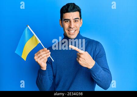 Handsome hispanic man wearing ukraine flag smiling happy pointing with ...