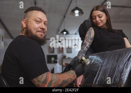 Happy bearded adult male tattooist in casual clothes sitting on stool ...