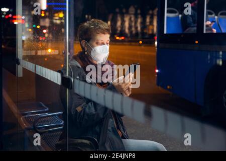 Woman sitting at a bus stop at night, looking at her phone with city ...