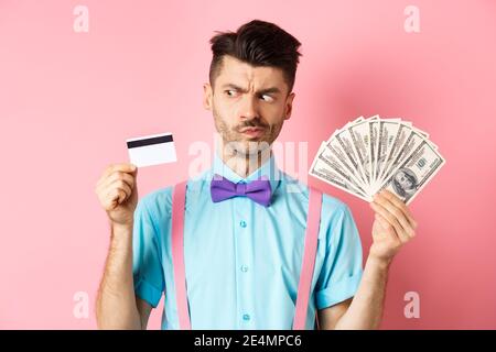 Doubtful and indecisive guy looking hesitant at plastic credit card ...