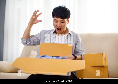 Smiling Asian guy unpacking online shopping at home, holding hoodie ...