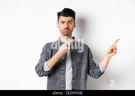 Sad caucasian guy pointing fingers right and pouting, looking hesitant ...