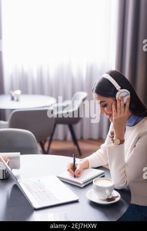 freelancer in headphones writing in notebook near laptop and smartphone ...