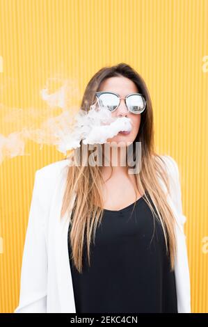 Woman exhaling smoke while standing against a brick wall in sunlight ...