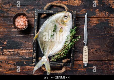Raw fish butterfish or pompano with herbs in a wooden tray. Dark wooden ...