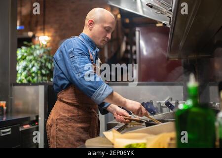 Bald chef in apron stirring dish on frying pan while cooking food in ...