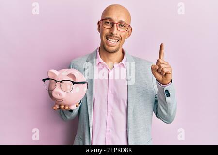 Bald man with beard holding piggy bank with glasses smiling with an ...