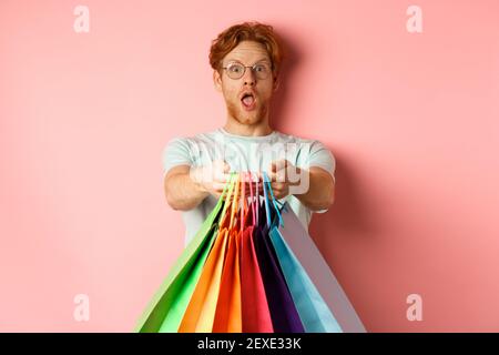 Surprised redhead man stretch out hands with shopping bags, give you ...