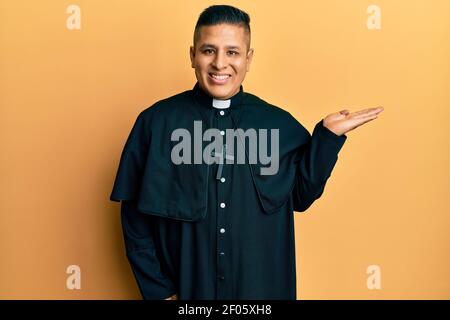 Young latin priest man standing over yellow background clapping and ...