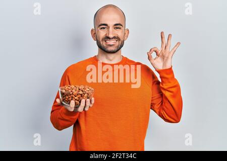 Young hispanic man holding peanuts doing ok sign with fingers, smiling ...