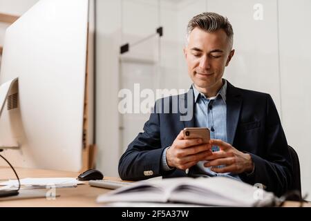 Pleased grey man using mobile phone while working with computer in ...