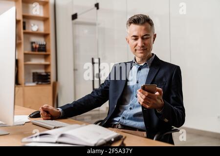 Pleased grey man using mobile phone while working with computer in ...