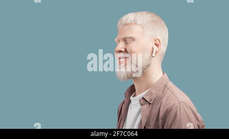 Relaxed albino guy listening music in bluetooth earpods, standing with ...