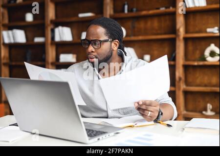 Focused biracial man reading paper letter working at laptop Stock Photo ...