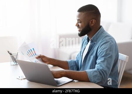 Black man working with laptop and papers while sitting on bench in park ...