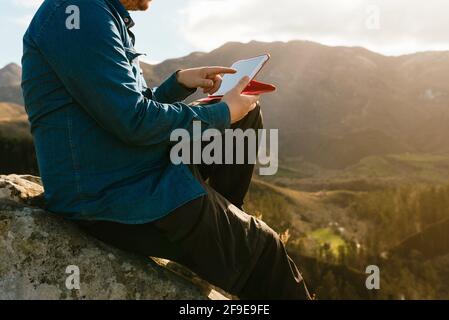 Tranquil male explorer with tablet sitting on hill in mountainous ...