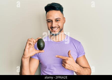 Young arab man holding avocado pointing thumb up to the side smiling ...