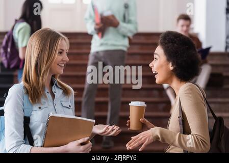 Positive multicultural students with notebook and coffee looking at ...