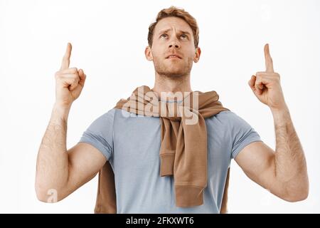 Confused young man frowning, pointing fingers up and looking displeased ...