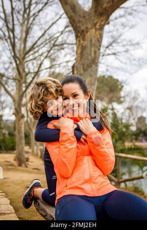 Charming boy embracing smiling mother while sitting in a wooden bench ...