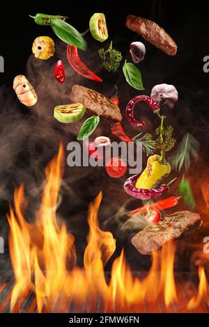 Flying beef stakes and vegetables above barbecue grill with fire on ...