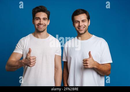 Joyful handsome guys smiling and showing thumbs up isolated over blue ...
