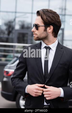 side view of bearded bodyguard in suit and sunglasses using walkie ...