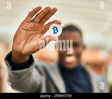 Young afro american man with bitcoin in hand in front of laptop in cafe ...