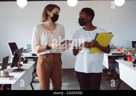 Two diverse businesswomen wearing face masks, holding tablet and ...