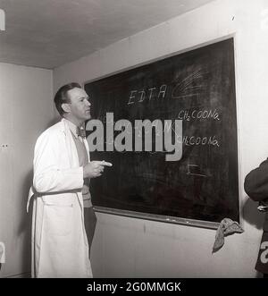 1950s SCHOOL TEACHER STANDING IN FRONT OF BLACKBOARD LEANING ON CHAIR ...