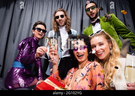 smiling interracial friends in colorful clothes drinking champagne near ...
