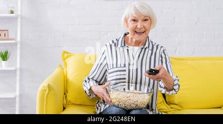 Elderly senior woman with remote controller in hands sitting on sofa in ...