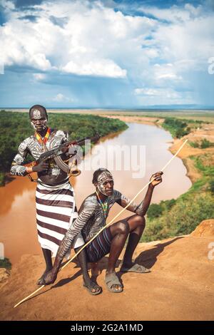 Men with a machine gun standing and sitting on cliff near dirty river ...