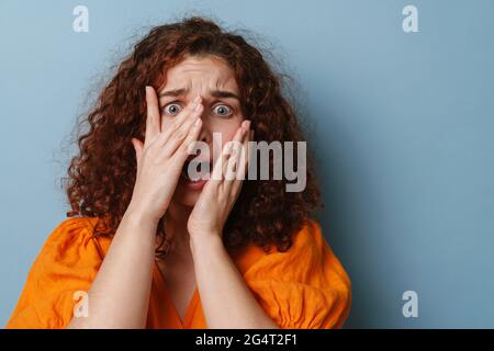 Scared ginger woman grimacing and covering her face isolated over blue ...