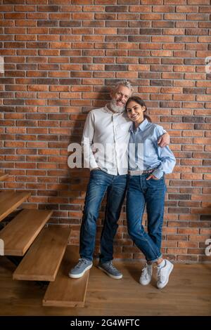 A gray-haired man hugging his wife while standing near the brick wall ...