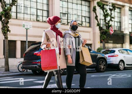 Muslim female friends in masks and with paper bags crossing road while ...