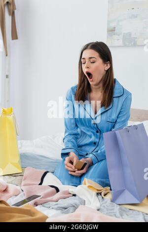 tired woman in stylish pajama yawning near shopping bags and assortment ...