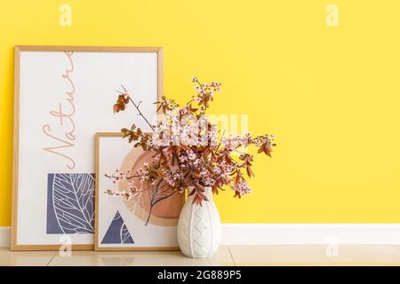 Vase with blossoming branches and pictures on floor near color wall ...