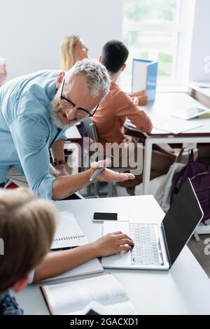 Mature teacher pointing with hand at notebook near smiling multiethnic ...