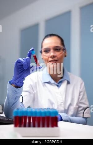 Biochemistry doctor analyzing vacutainer with blood while having microscope on desk and using ...