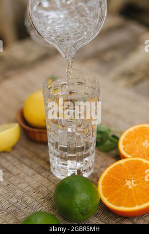 Proces of pouring cold water into a tall glass. Fresh citrus on table ...