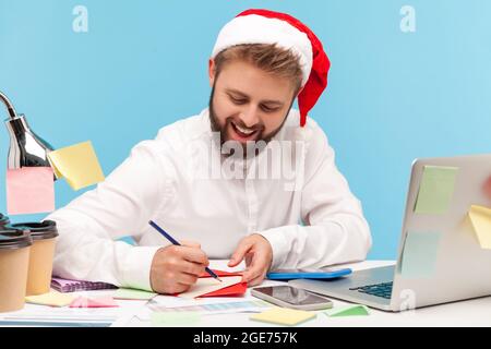 Happy bearded Santa Claus writing on wish list sitting home at workshop ...