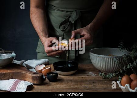 Unrecognizable guy in apron breaking raw egg over bowl while preparing ...