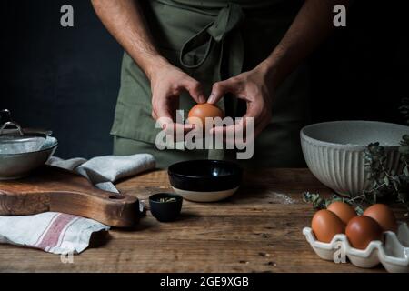 Unrecognizable guy in apron breaking raw egg over bowl while preparing ...