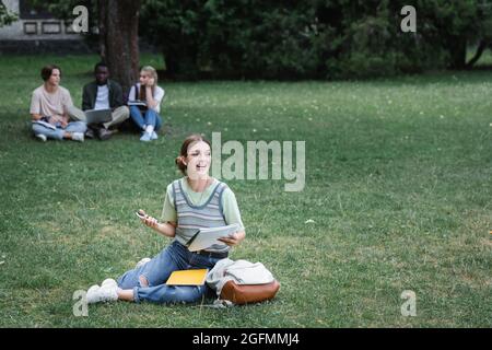 Cheerful interracial students with notebook and smartphone standing ...