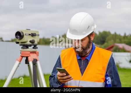 A civil engineer with an optical level looks into a mobile phone. A ...