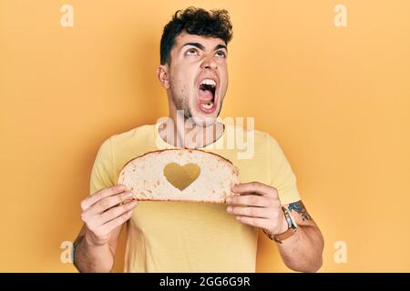 Young hispanic man holding bread dough and cooking tools angry and mad ...