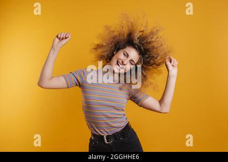 Happy excited young casual woman with fizzy hairstyle standing over yellow wall background ...